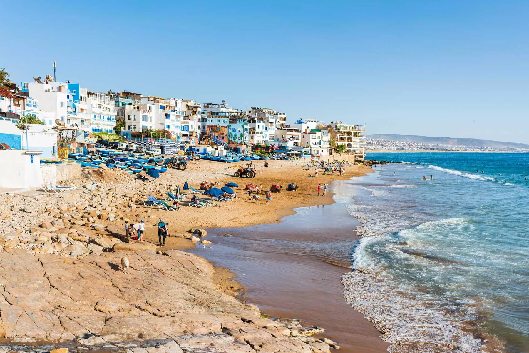 Muonekano wa ufuo wa Taghazout, Morocco mchana wa jua sana na wapenda ufuo kuonekana wakitembea ufukweni, wakiota jua au kuogelea.