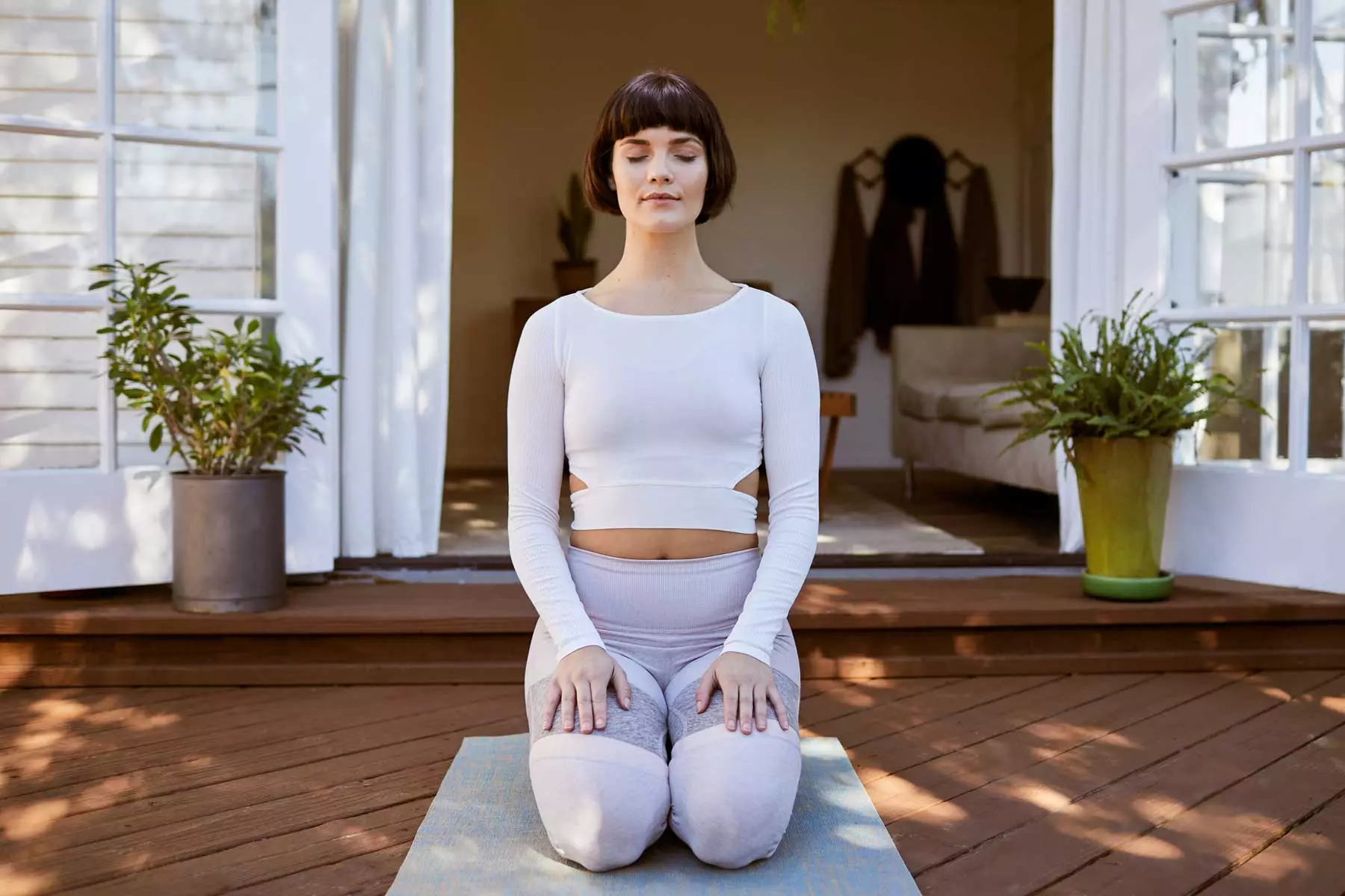 Wanita muda dalam pakaian sukan melutut di atas tikar senaman di teresnya dan bermeditasi dengan mata tertutup semasa meditasi