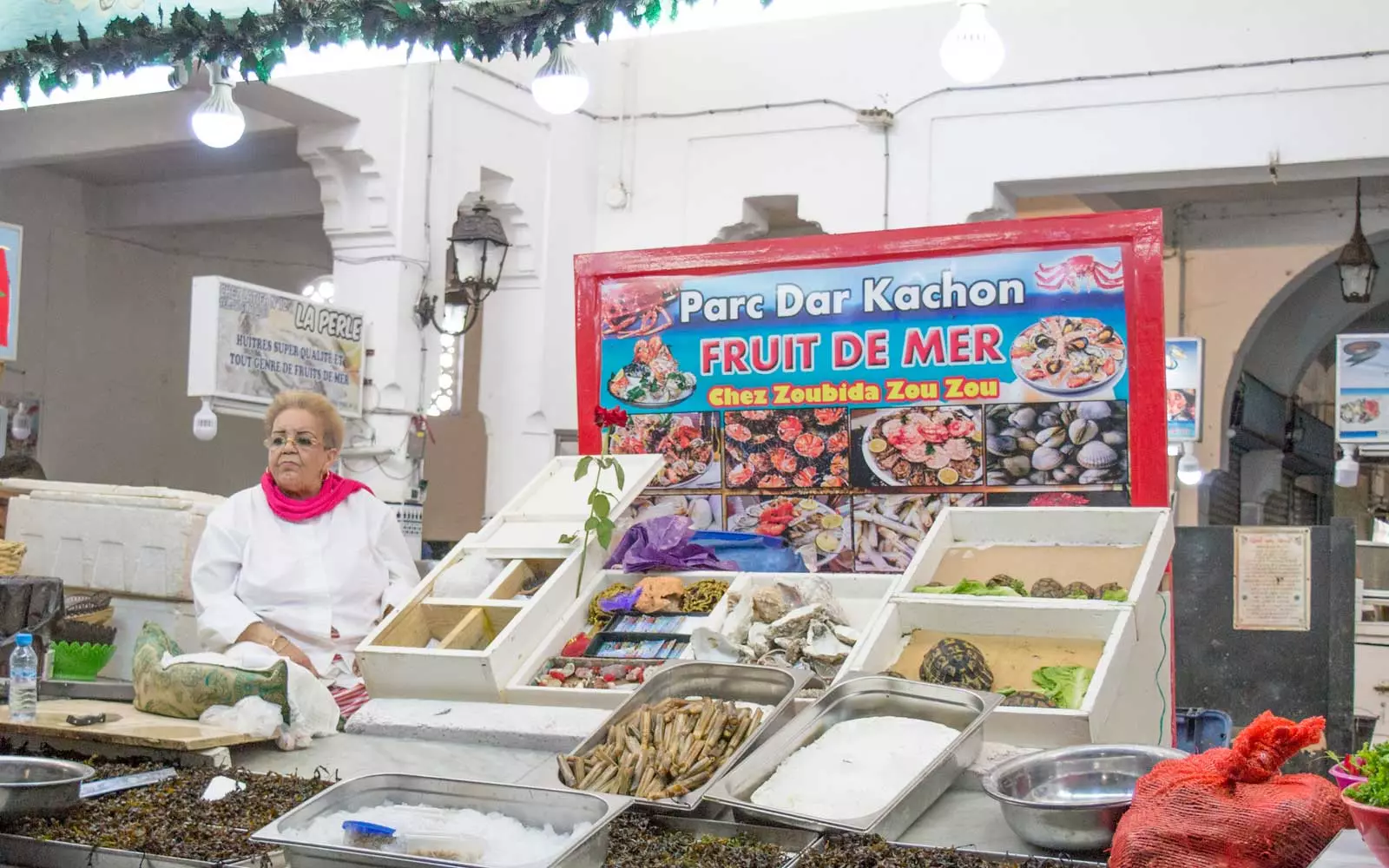 Vrouw verkoper chats met een mannelijke klant. Er zijn bakjes met zeewier, kokkels en krab te zien. Op de achtergrond zijn foto's van zeevruchten, waaronder mosselen en langoustine plus een bord van de stand met 'Seafood at Zoubida Zou Zou's