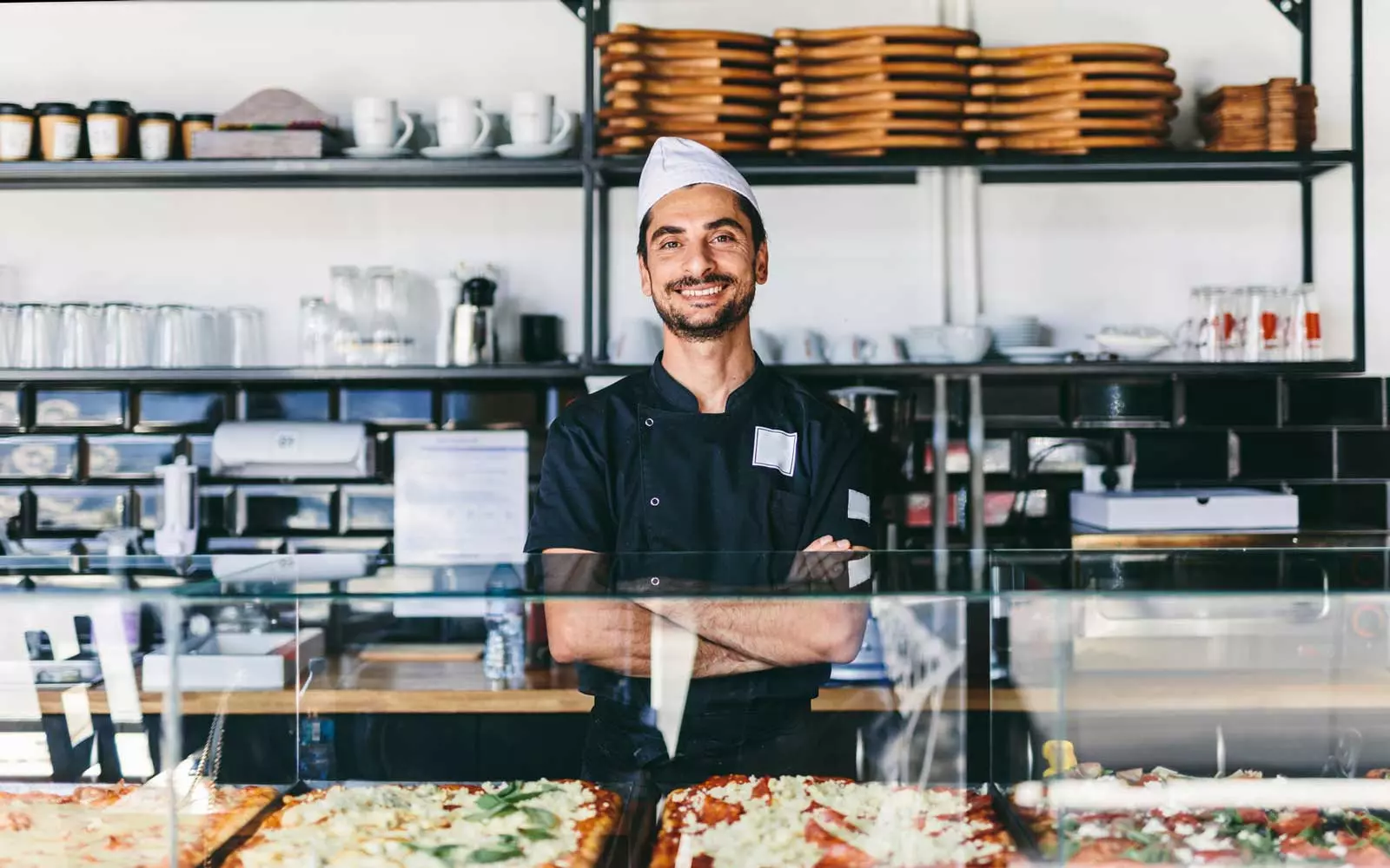 Hoton mai dafa abinci mai murmushi tare da ketare hannaye tsaye ta wurin nunin dillali a cikin pizzeria