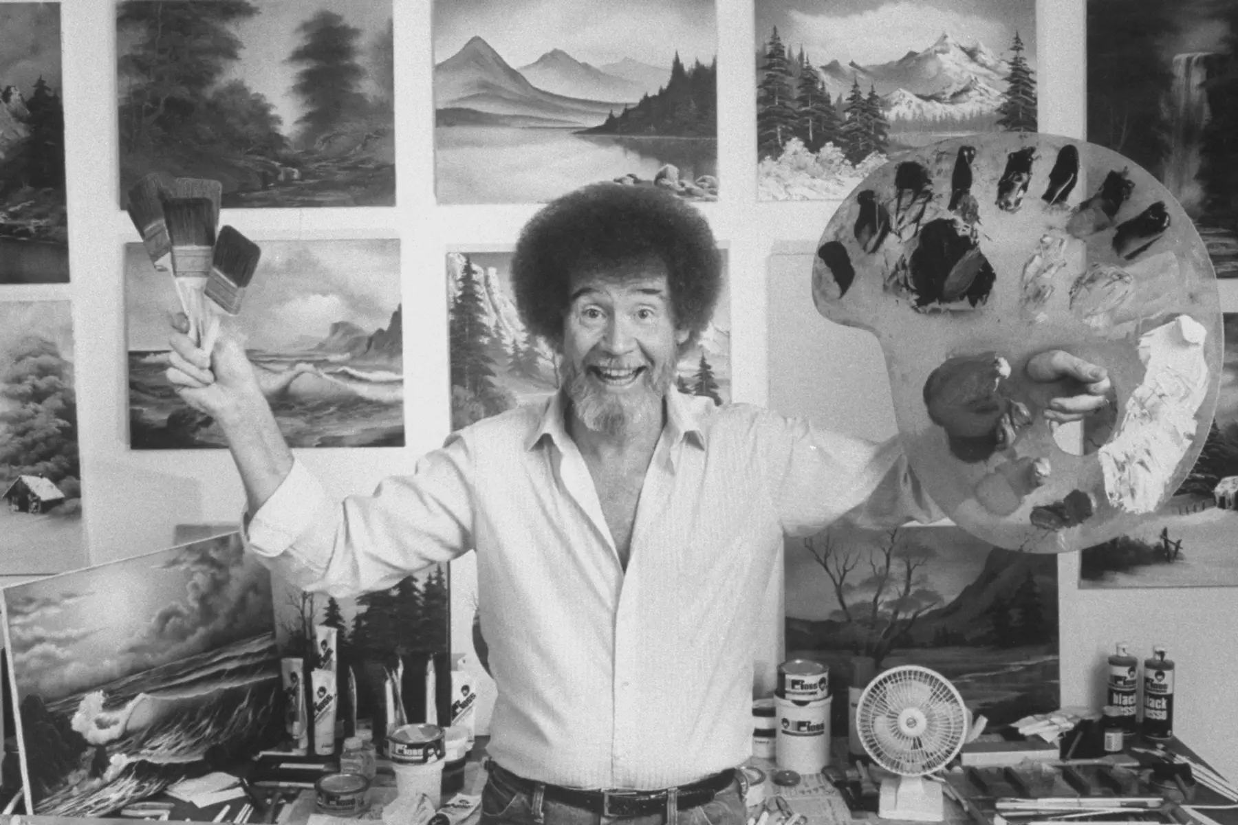 L'instructor/artista de pintura de televisió Bob Ross aixeca alegrement la paleta de pintura i els pinzells mentre es troba davant de la paret coberta. les seves pintures de paisatge al seu estudi.