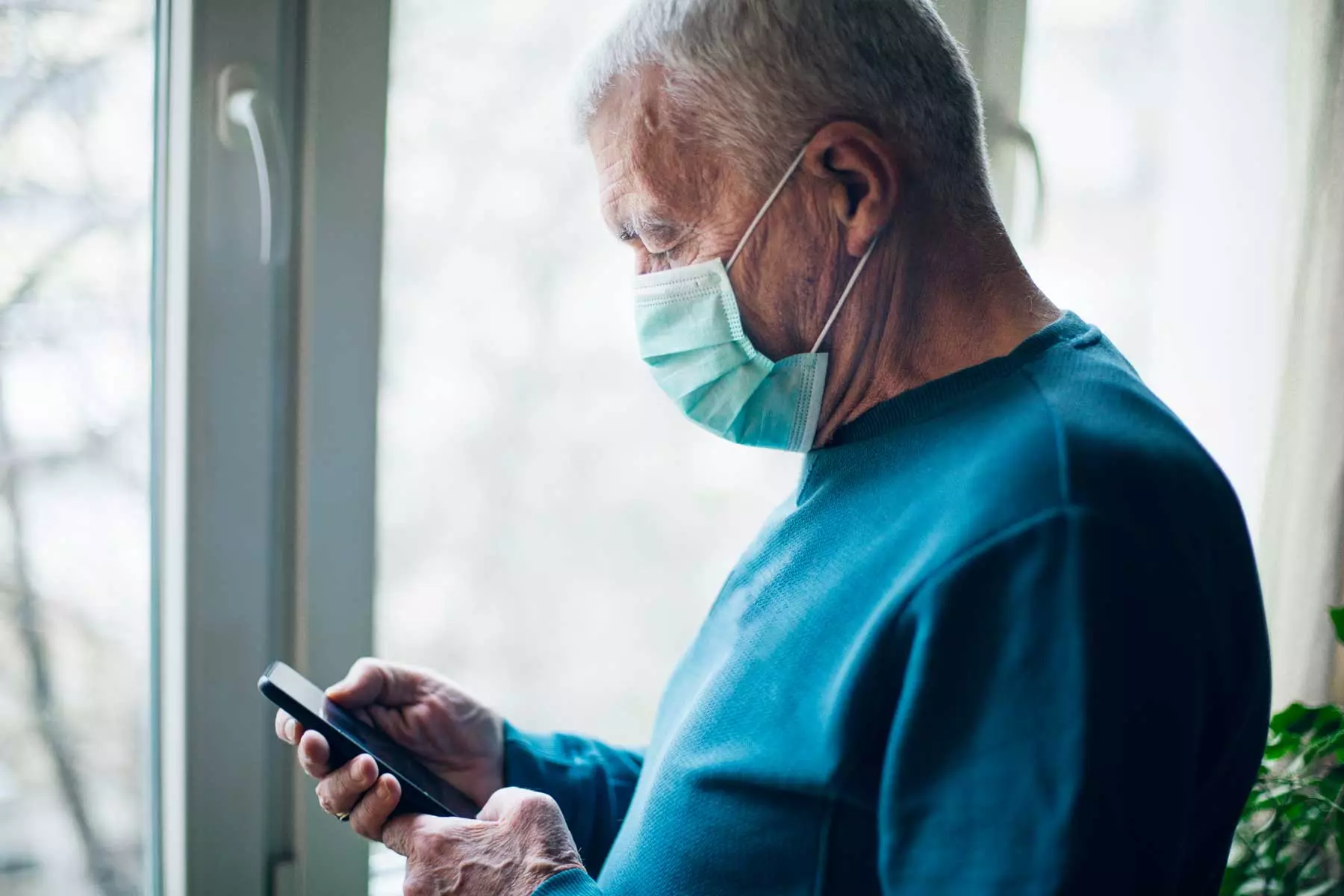 Home gran que porta una màscara protectora i utilitza un telèfon a casa durant la pandèmia de la COVID-19.
