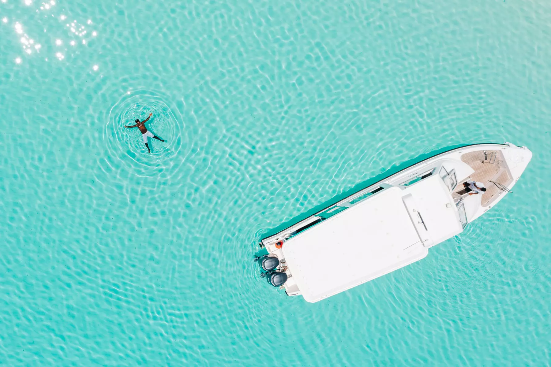क्रिस्टल-स्वच्छ निळ्या पाण्यात बोटीजवळ तरंगणाऱ्या व्यक्तीचा हवाई फोटो