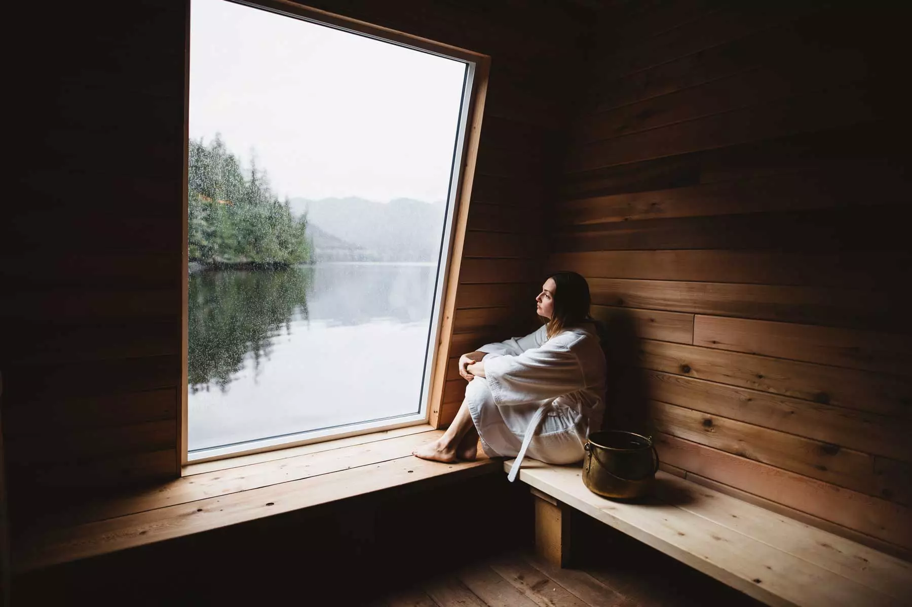 Donna seduta nella sauna che fissa fuori dalla finestra l'acqua e gli alberi