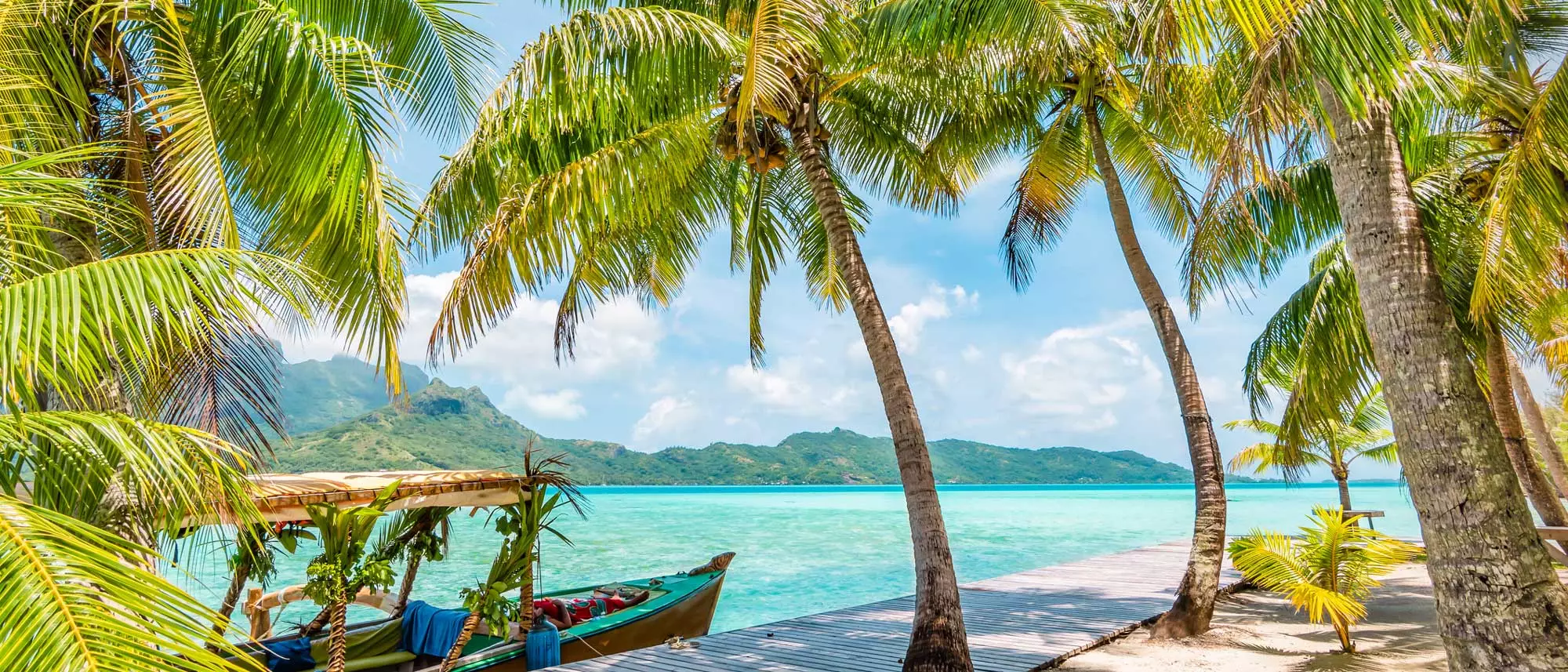 Vackert sommarlandskap med kokospalmer på den tropiska ön Bora Bora, Franska Polynesien. Dekorerad turistbåt förtöjd vid träkajen.
