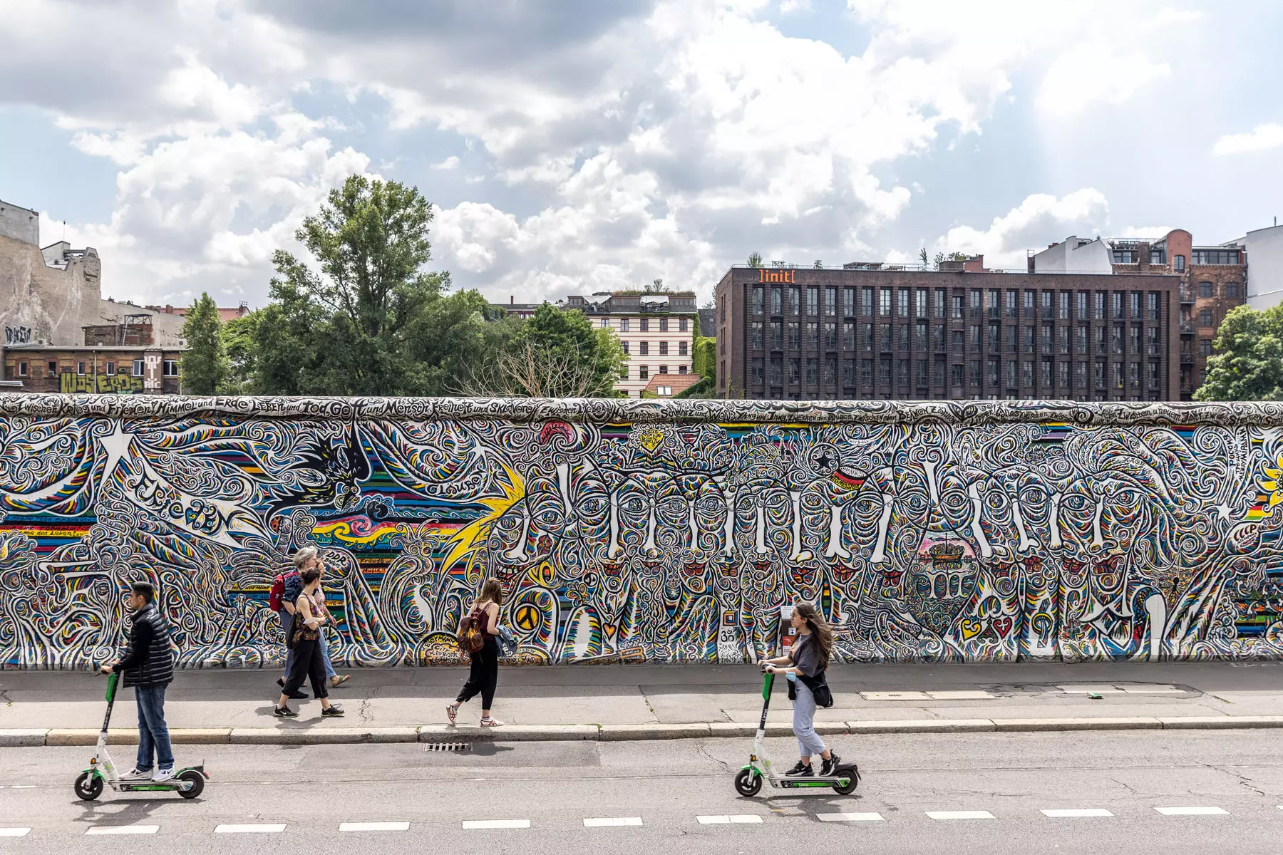 Lidé jezdí na koloběžkách poblíž East Side Gallery, galerie pod širým nebem na nejdelší dochované části Berlínské zdi, 3. července 2021 v Berlíně v Německu.