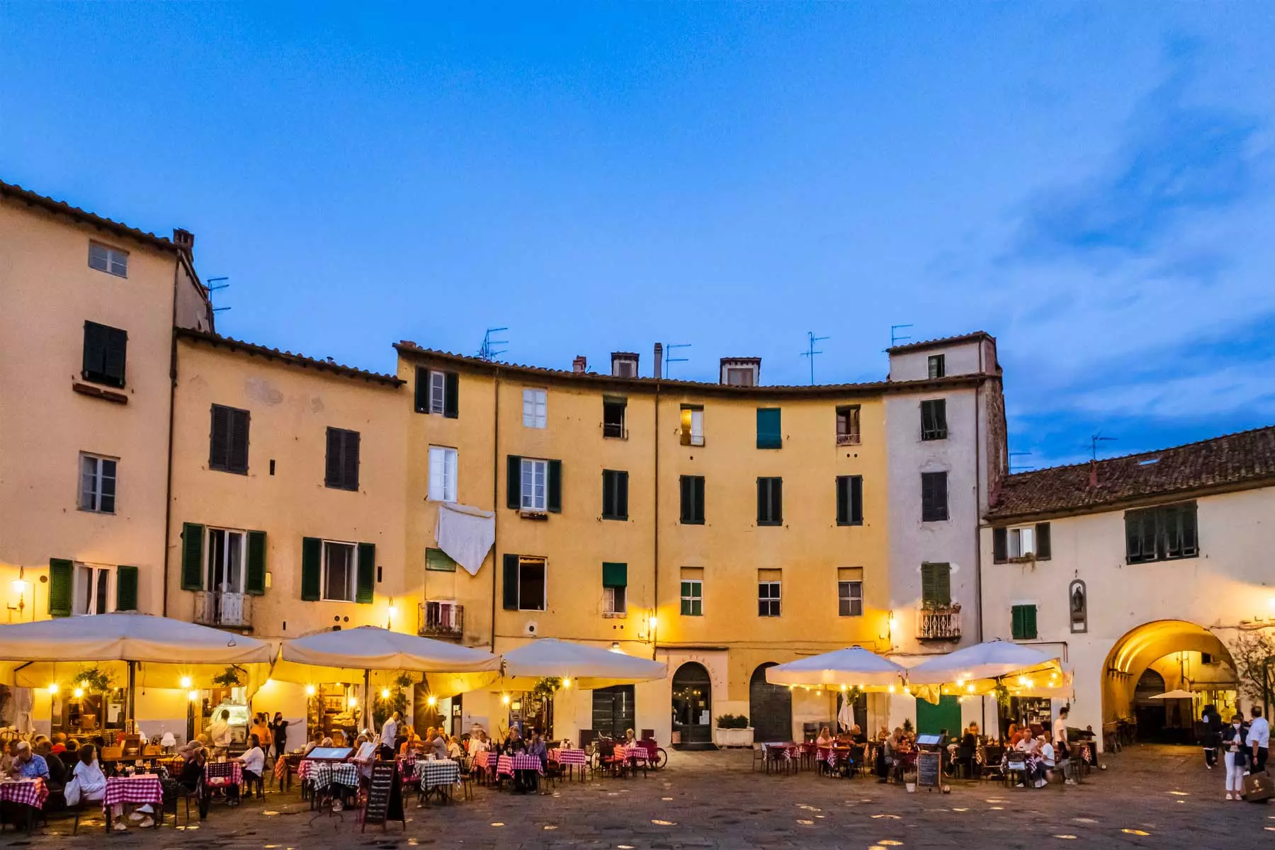 Turis duduk di meja luar ruangan di Piazza dell'Anfiteatro of Lucca, alun-alun berbentuk elips yang dikelilingi oleh cincin bangunan yang mengikuti bekas amfiteater Romawi abad kedua di kota.