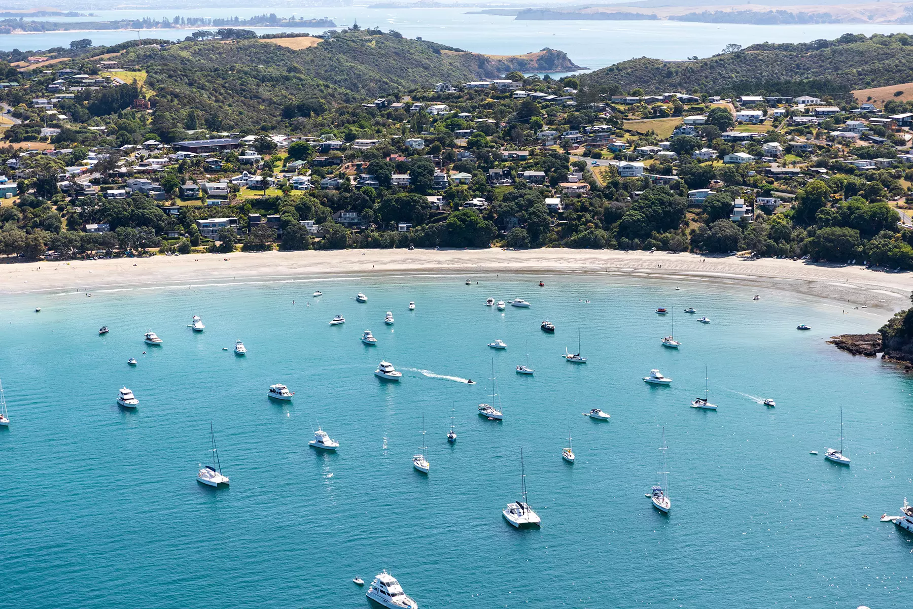 vue aérienne de l'île de Waiheke avec du sable, des arbres et des maisons sur le rivage et des bateaux dans l'océan environnant