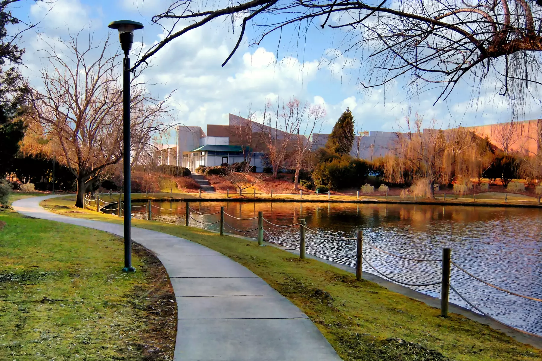 Šetnja uz jezero u University Villageu, Charlotte, Sjeverna Karolina na vedri, topli zimski dan. Drveće je golo, ali sunce sija kroz vedro nebo s oblacima, a svjetlost je zlatna.