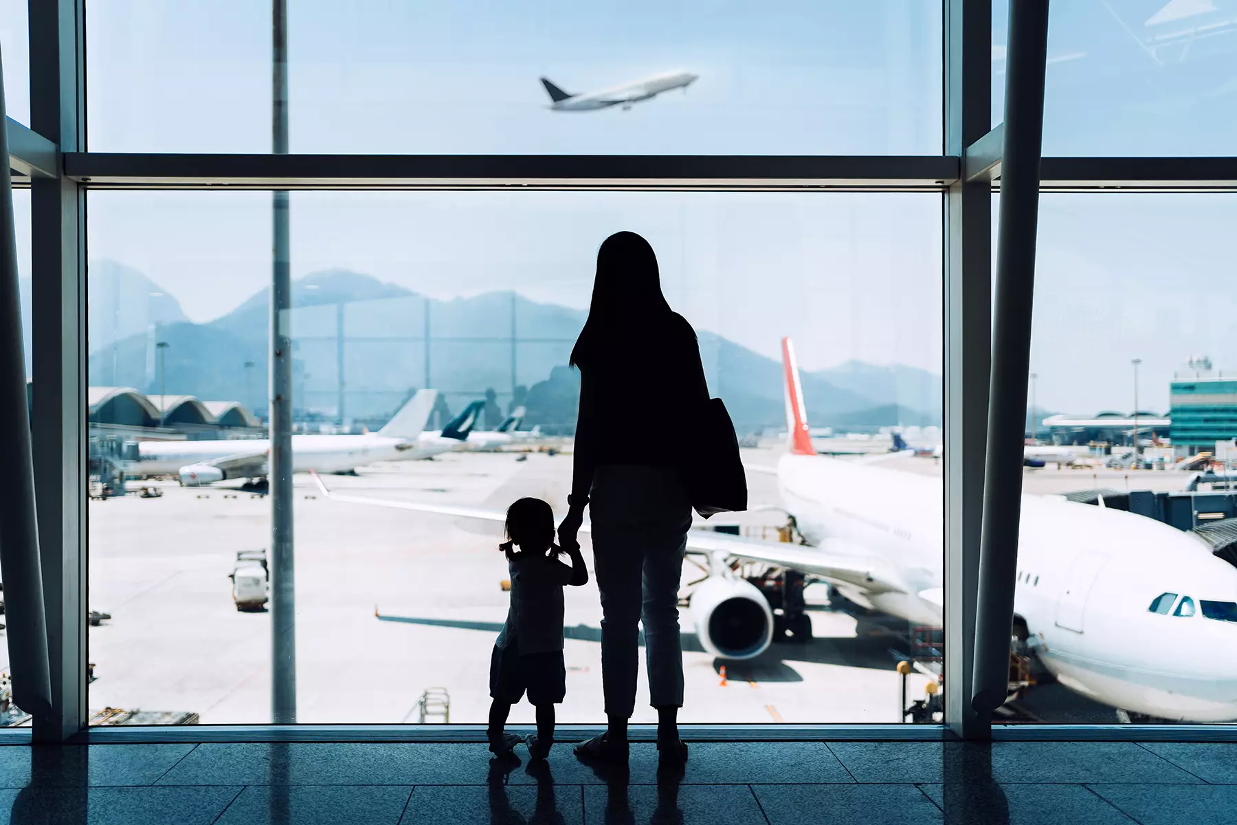 Seorang ibu memegang tangan anak perempuan melihat kapal terbang melalui tingkap di lapangan terbang sementara menunggu berlepas
