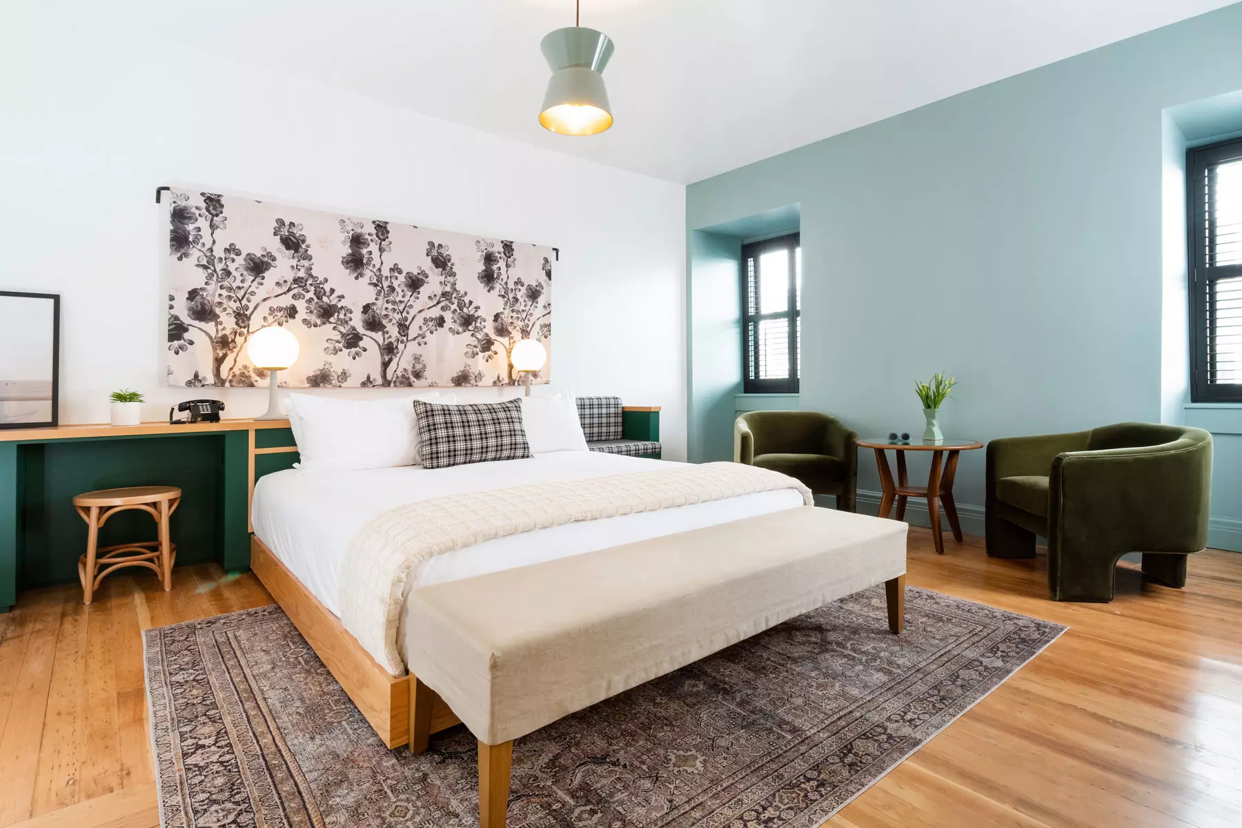 голема хотелска соба со кревет со цветни шари, зелена маса и фотелји за фотелји и воздушна соба исполнета со сонце
