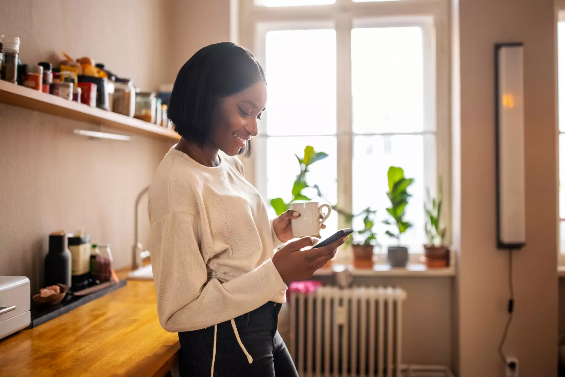 Wanita berdiri di dapur memegang secawan kopi dan menghantar mesej di telefon bimbitnya. Wanita menggunakan telefon bimbit di rumah.