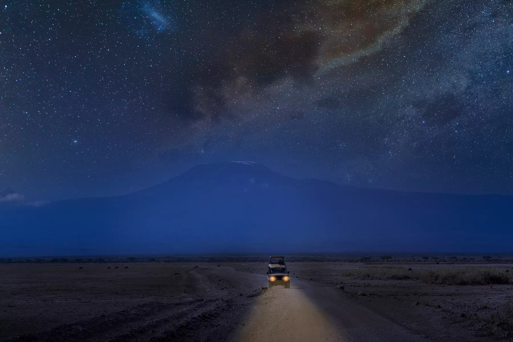 Tampilan depan kendaraan berpenggerak empat roda di jalan tanah di bawah Gunung Kilimanjaro di malam berbintang dengan Bima Sakti.