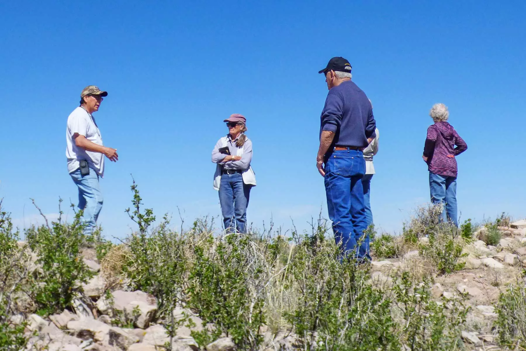 Fënnef Leit stinn op Fielsen a Gras mat engem bloen Himmel hannendrun, zu Zuni Pueblo