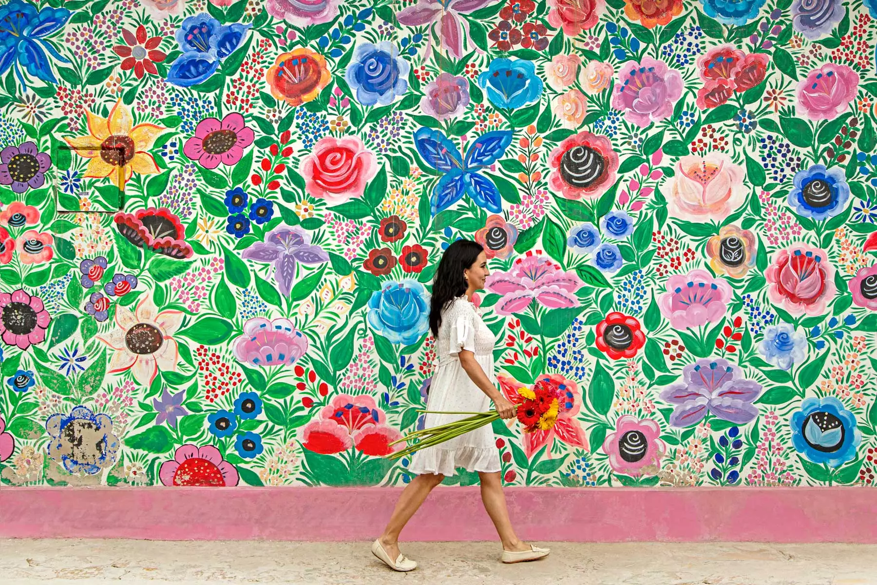 白いドレスを着た女性がメキシコの色とりどりの花の壁画を通り過ぎて歩く