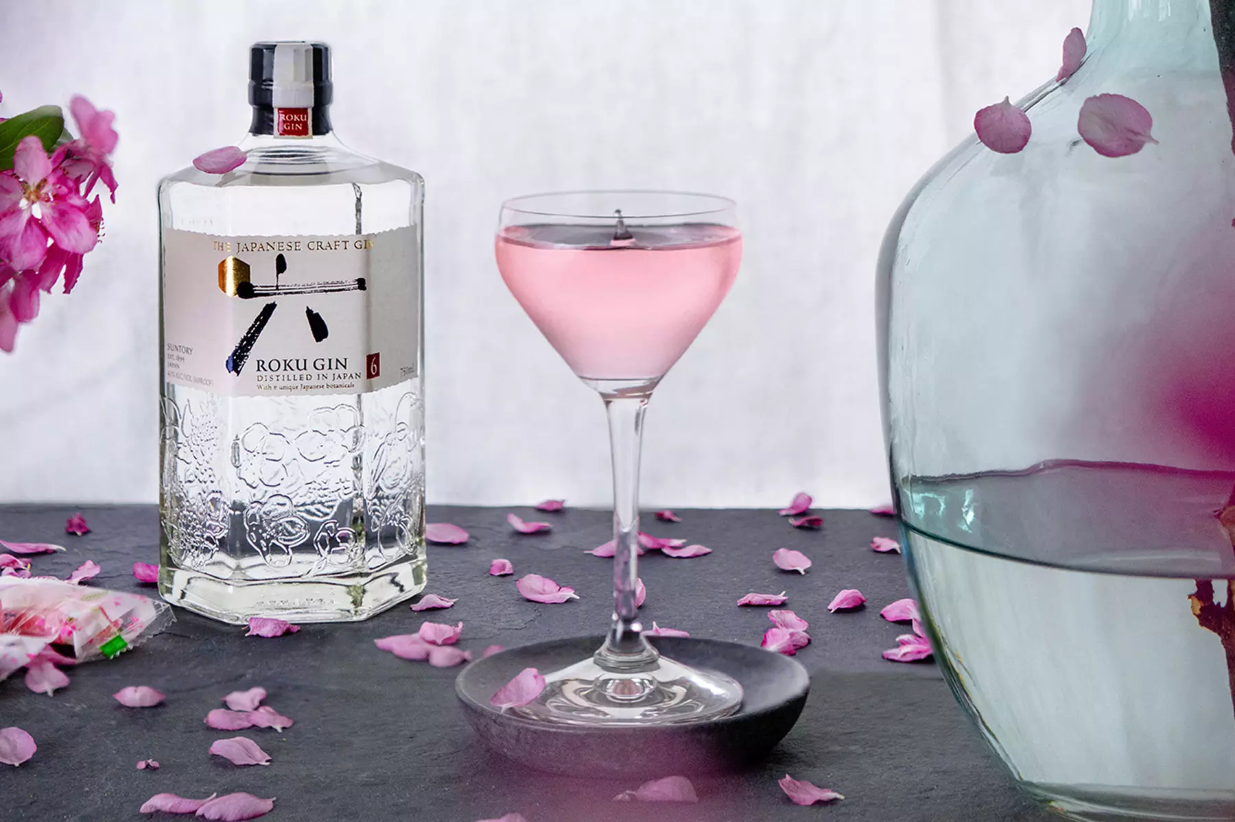 mmanya pink na iko coupe n'akụkụ karama Roku Craft gin, ifuru cherry gbasasịrị na tebụl.