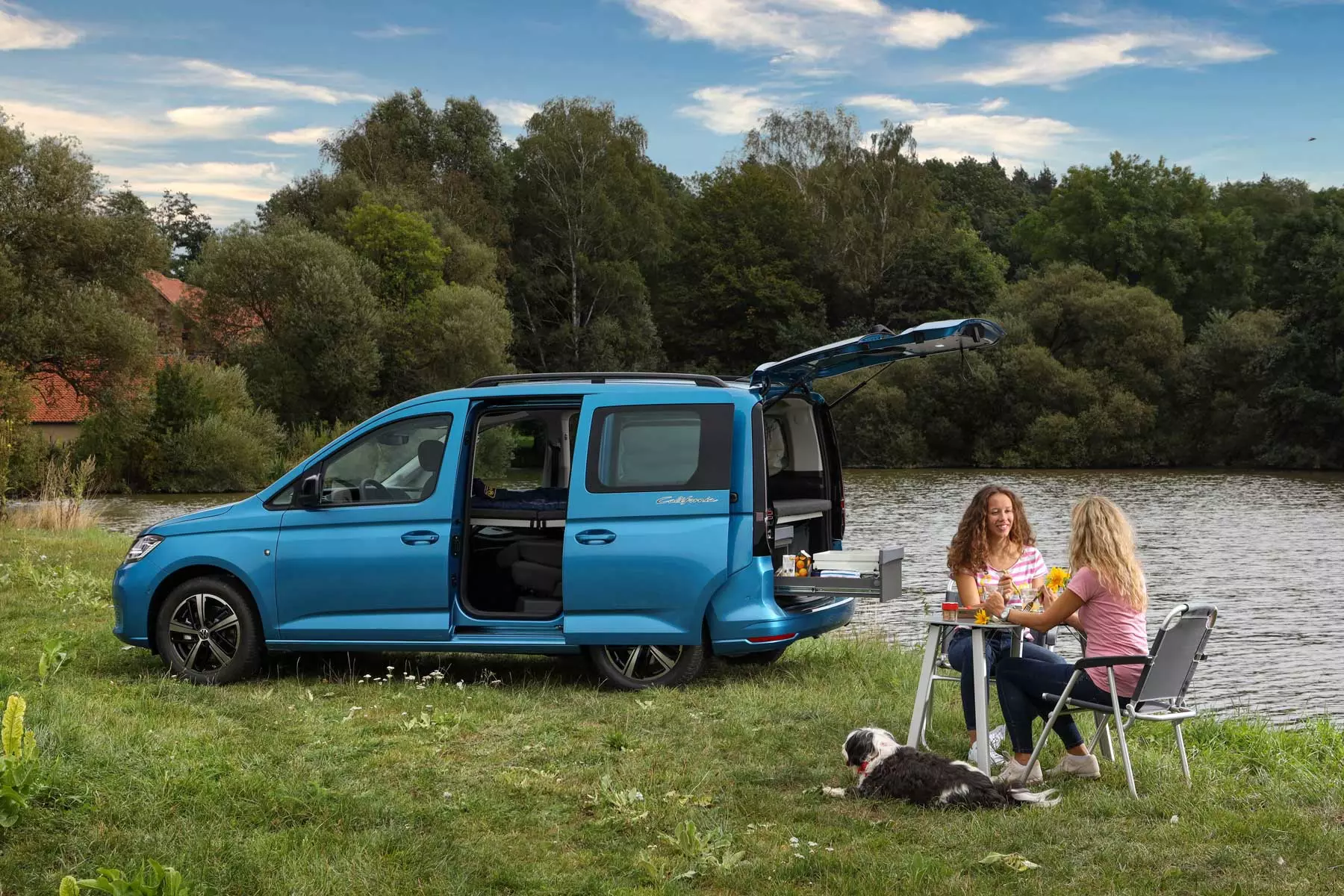 Két nő táboroz a folyó közelében a 2021-es Volkswagen California Caddyben a kutyájával