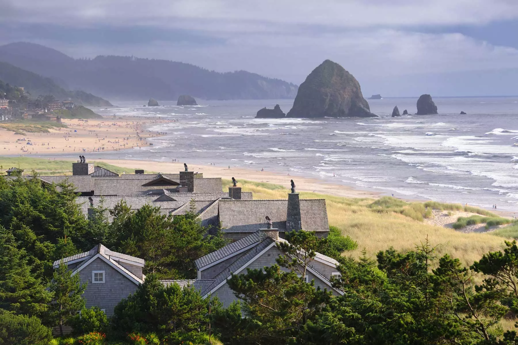 Slávna skala Haystack Rock s neuveriteľnou oblačnosťou, ktorá sa tiahne na obzore. Hmla vĺn sa rozprašuje spredu, zatiaľ čo okolo prírodnej rezervácie poletujú všetky druhy vtákov. Veľa ľudí dole na pláži si užíva tento nádherný letný deň.