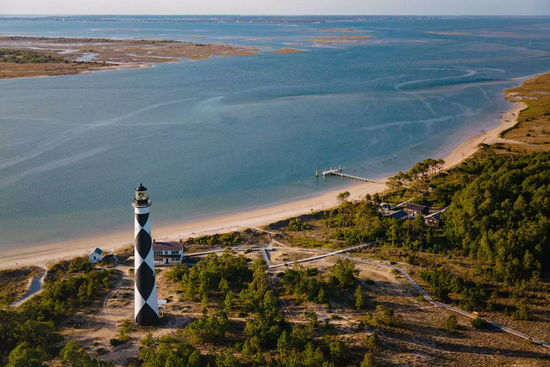 Aerial view ng Cape Lookout National Seashore, Cape Lookout Lighthouse at Keepers' Quarters Museum, mga opisina ng National Park Service, gift shop at dock na may Barden's Inlet, Shackleford Banks at Harker's Island sa di kalayuan at berdeng mga dahon sa baybayin.