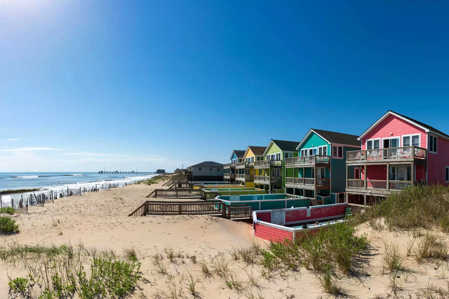 Side view ng hilera ng mga makukulay na beach house sa Nags Head. Pink na bahay simula sa hilera, navy house sa huling hanay. Buhangin at Beach sa kaliwa. Pier sa background. Mga tao sa pool sa pink na bahay.