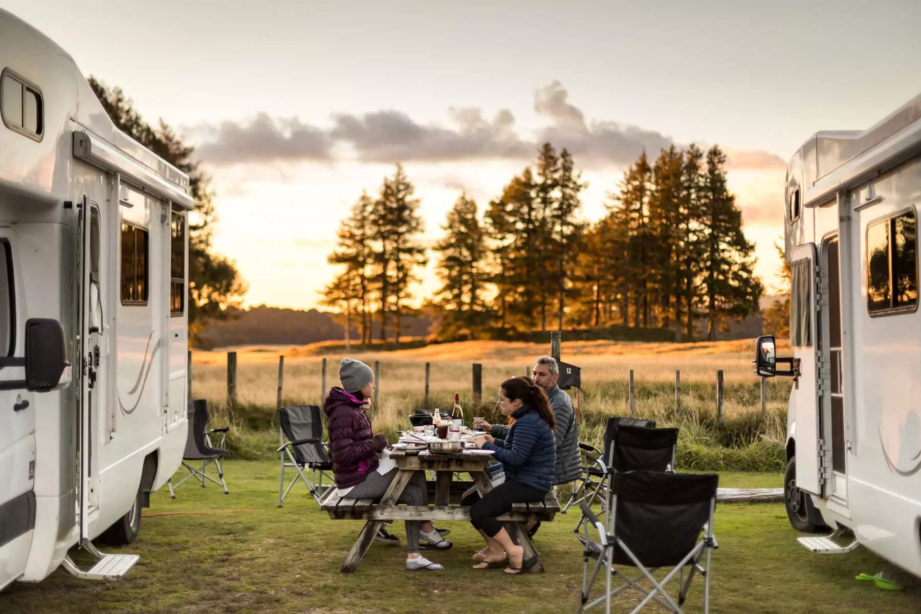 Мушкарац и две жене вечерају на отвореном око дрвеног стола за пикник док сунце залази