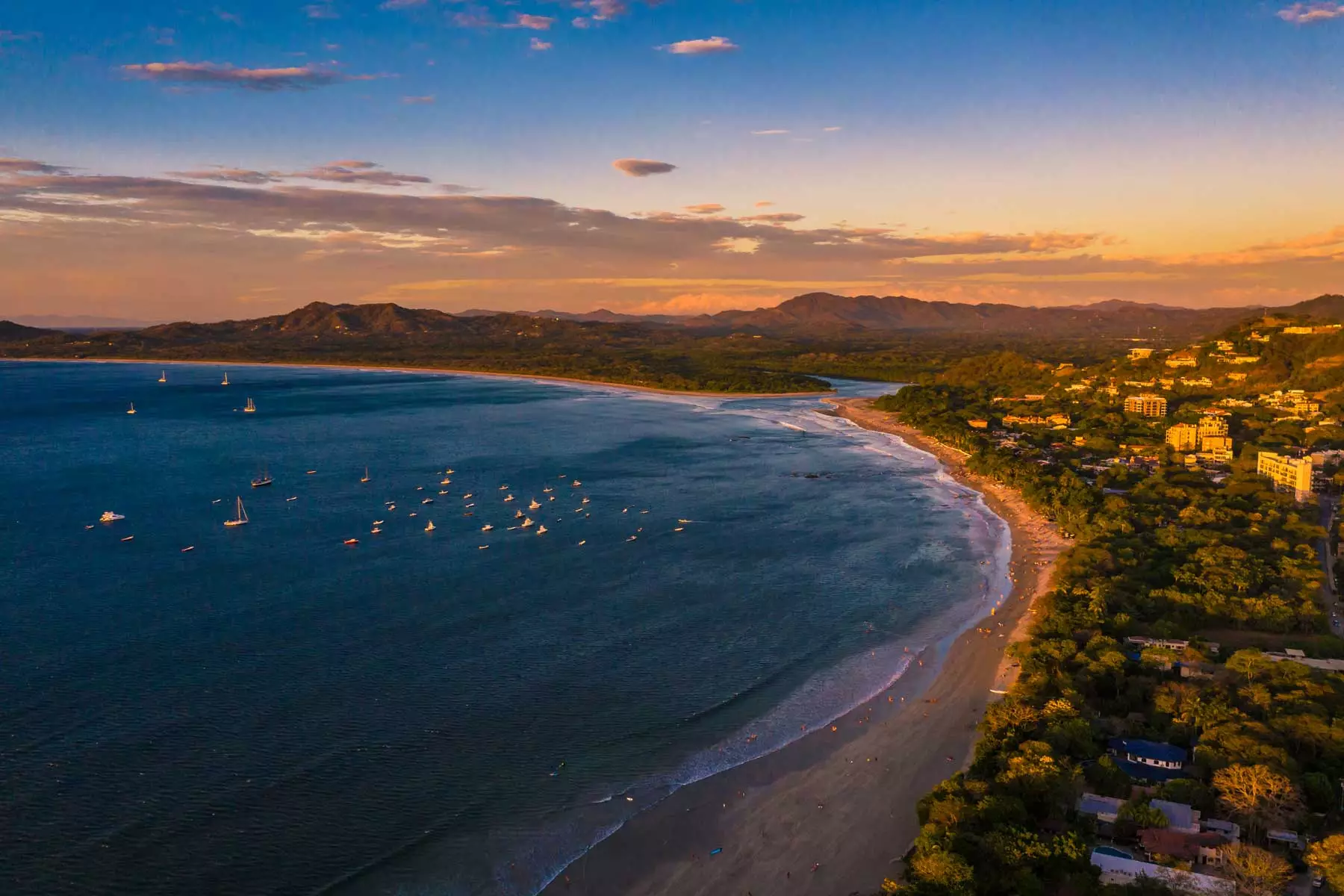 Mwonekano wa angani wa Tamarindo Beach katika mwanga wa jua wa chungwa. Unaweza kuona boti na majengo ya kando ya vilima kwenye mstari huu wa pwani ya Kosta Rika