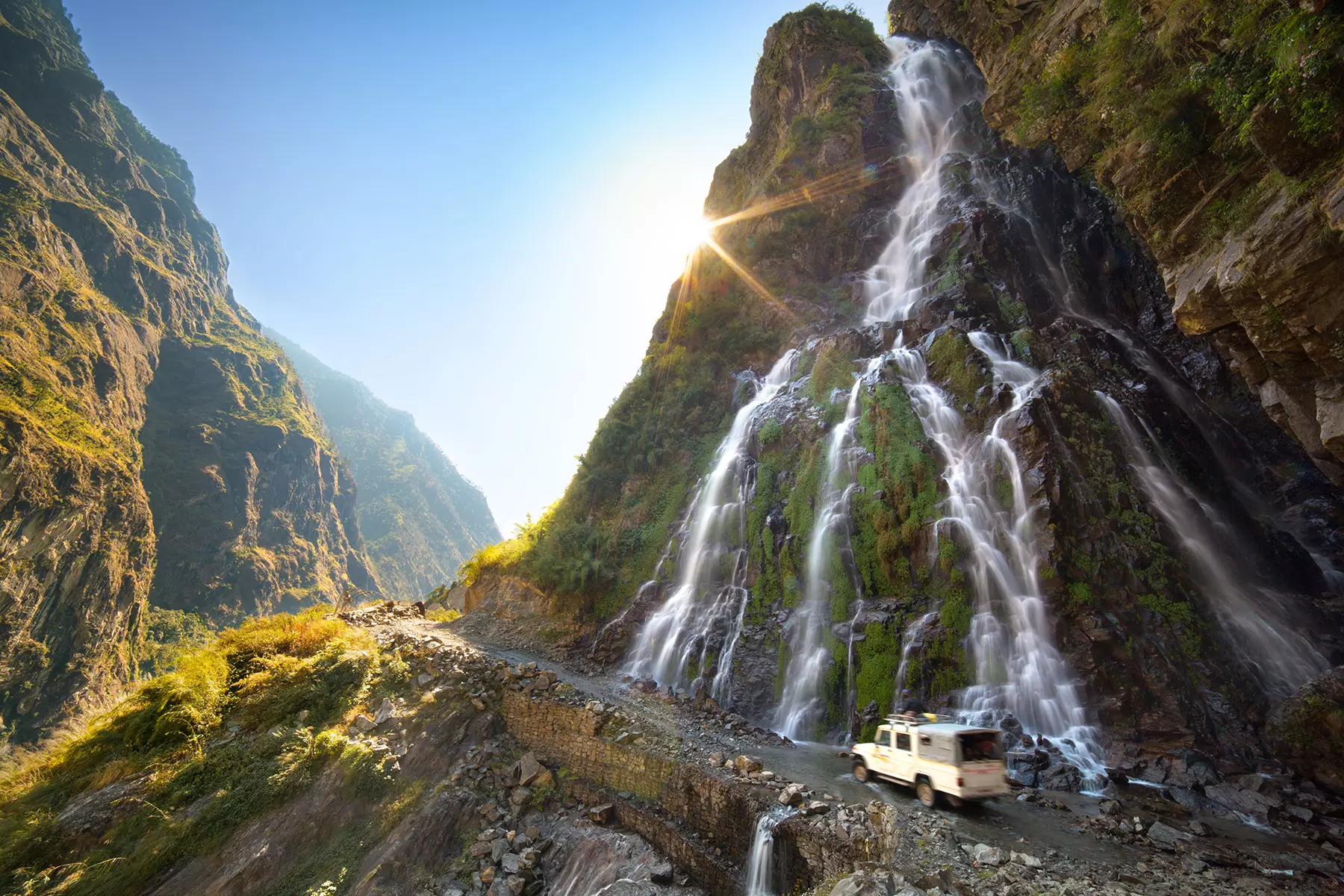 Naik mobil melintasi jalan pegunungan yang cerah di bawah air terjun di Nepal, Himalaya, Kawasan Konservasi Annapurna.