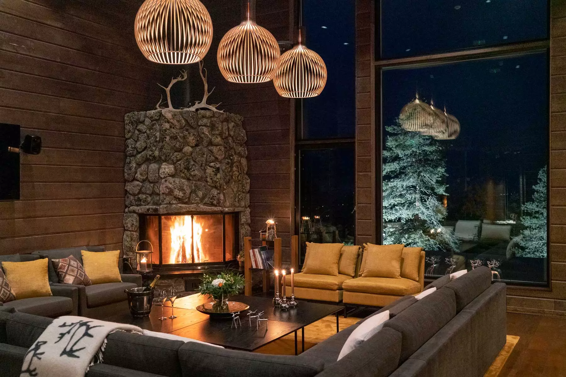 De lounge van de luxe lodge Octola in Finland, met een open haard met een knapperend haardvuur en een grote bank; bomen en de nachtelijke hemel zijn zichtbaar door het raam