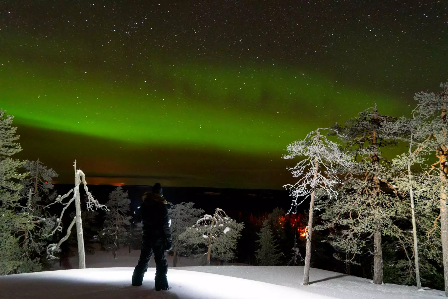 Mae dyn wedi'i wisgo mewn dillad gaeaf yn arsylwi'r Northern Lights o deras wedi'i orchuddio ag eira yn y Ffindir